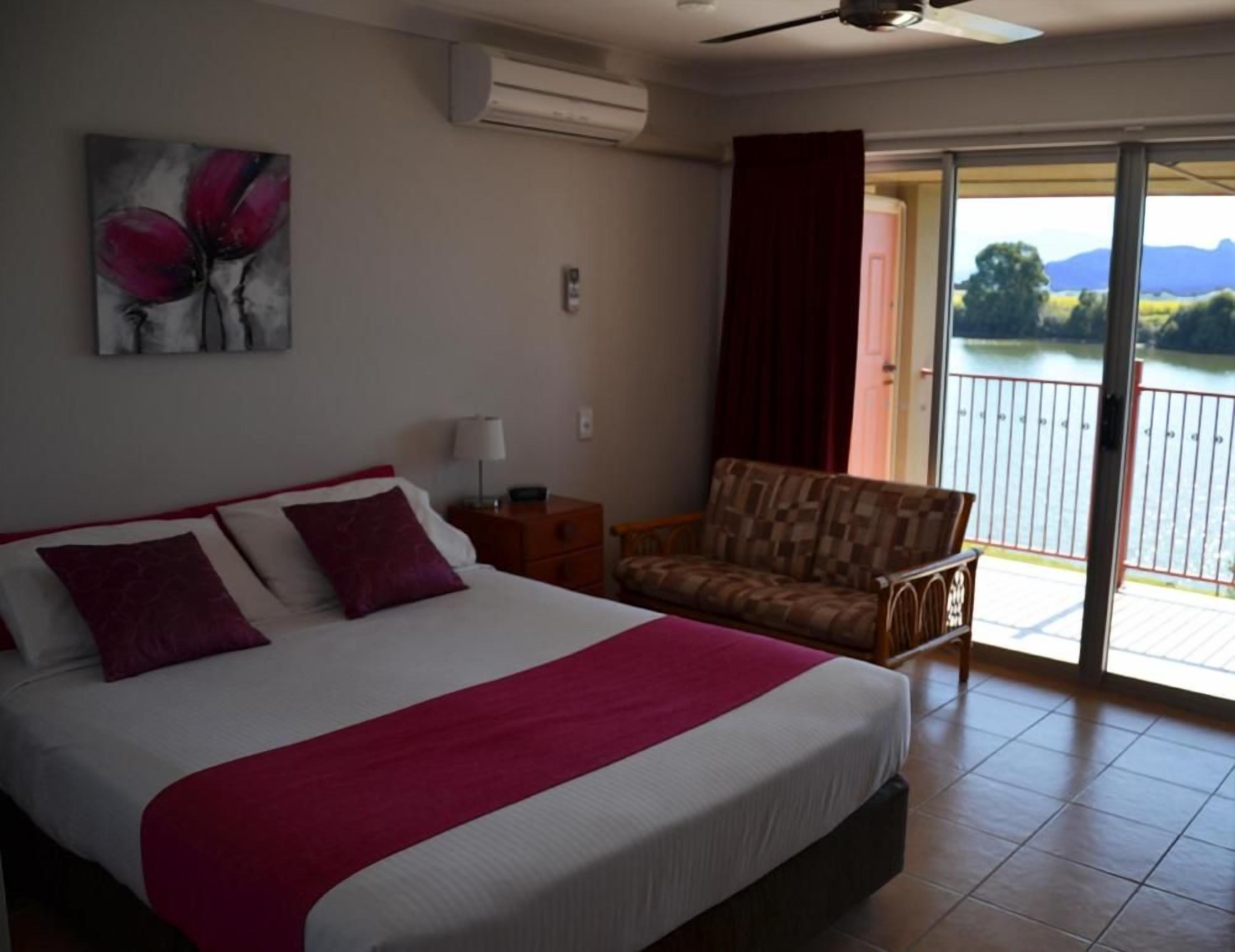 Quarto de hotel com uma cama grande, uma cadeira, um quadro e vista para um corpo de água.