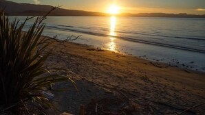 Uma praia com o pôr do sol sobre a água, uma faixa de areia e alguma vegetação ao primeiro plano.