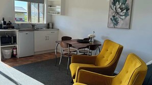 Cozinha moderna com armários brancos e uma janela, área de jantar com mesa e cadeiras, e uma poltrona amarela.