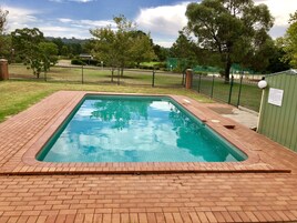 Piscina retangular com borda de tijolos, cercada por uma área com árvores e um campo gramado ao fundo.
