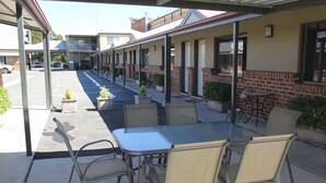 Uma área de estar coberta ao ar livre com mesas e cadeiras, um estacionamento e uma fileira de edifícios com fachada de tijolos e grandes janelas.
