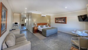 Quarto de hotel com cama, mesas de cabeceira, televisão, área de estar com sofá e cadeiras, e uma pequena mesa com copos e pratos.
