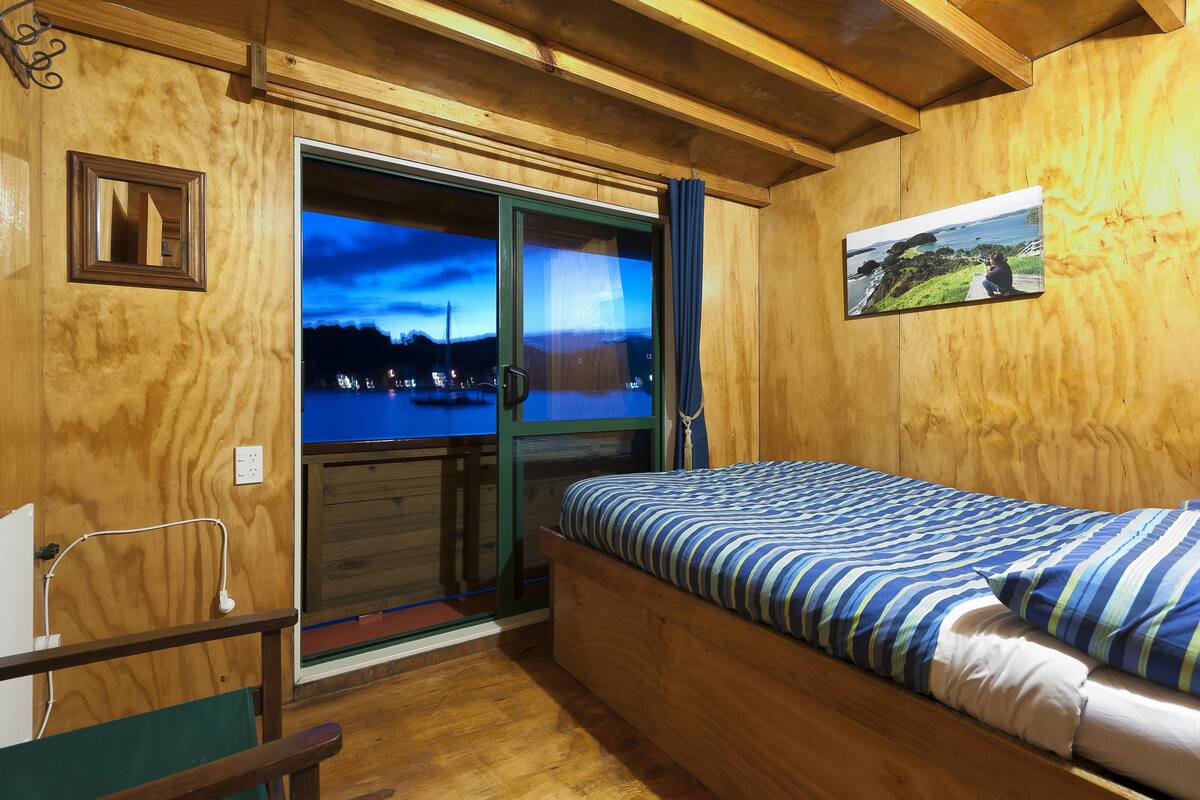 Quarto de madeira com uma cama, uma escrivaninha e janela com vista para o mar.