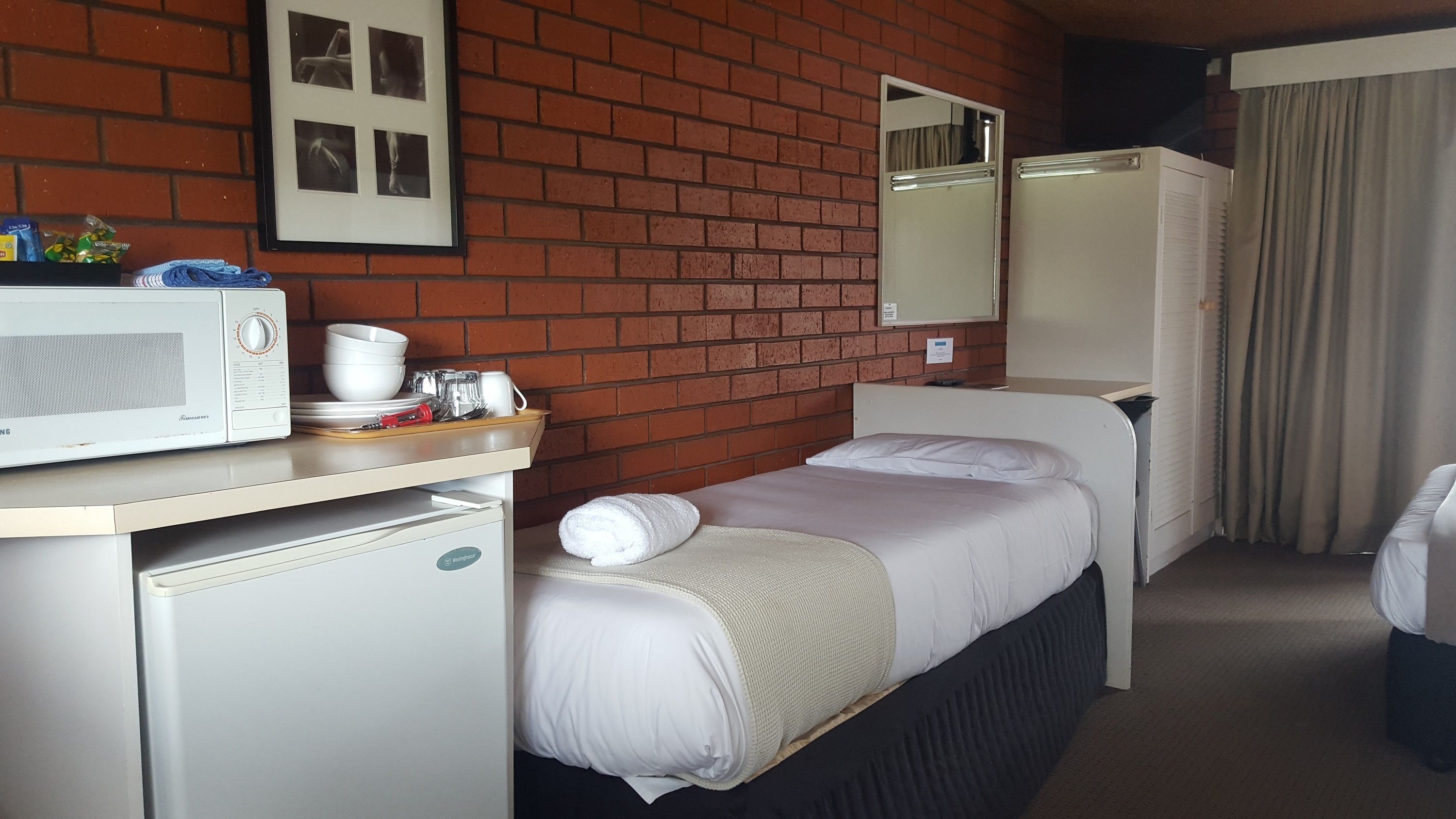 Quarto de hotel com parede de tijolos, cama com roupa de cama branca, pequena mesa com micro-ondas e espelho sobre a cama.