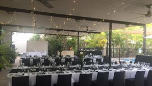 Uma área de refeições ao ar livre com mesas montadas para um evento formal, com luzes de corda e ventiladores de teto.