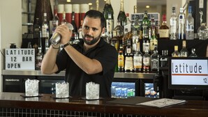 Um bartender em um bar, com diversas garrafas de bebidas alcoólicas ao fundo.