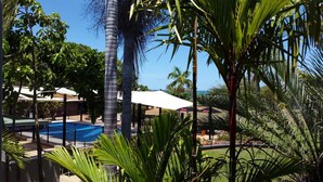 Área da piscina com vista clara para o oceano, cercada por palmeiras e um espaço de estar coberto.