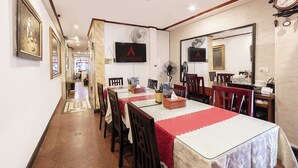 Uma sala de jantar de hotel com uma mesa comprida, cadeiras e uma tela de TV exibindo o nome do hotel.