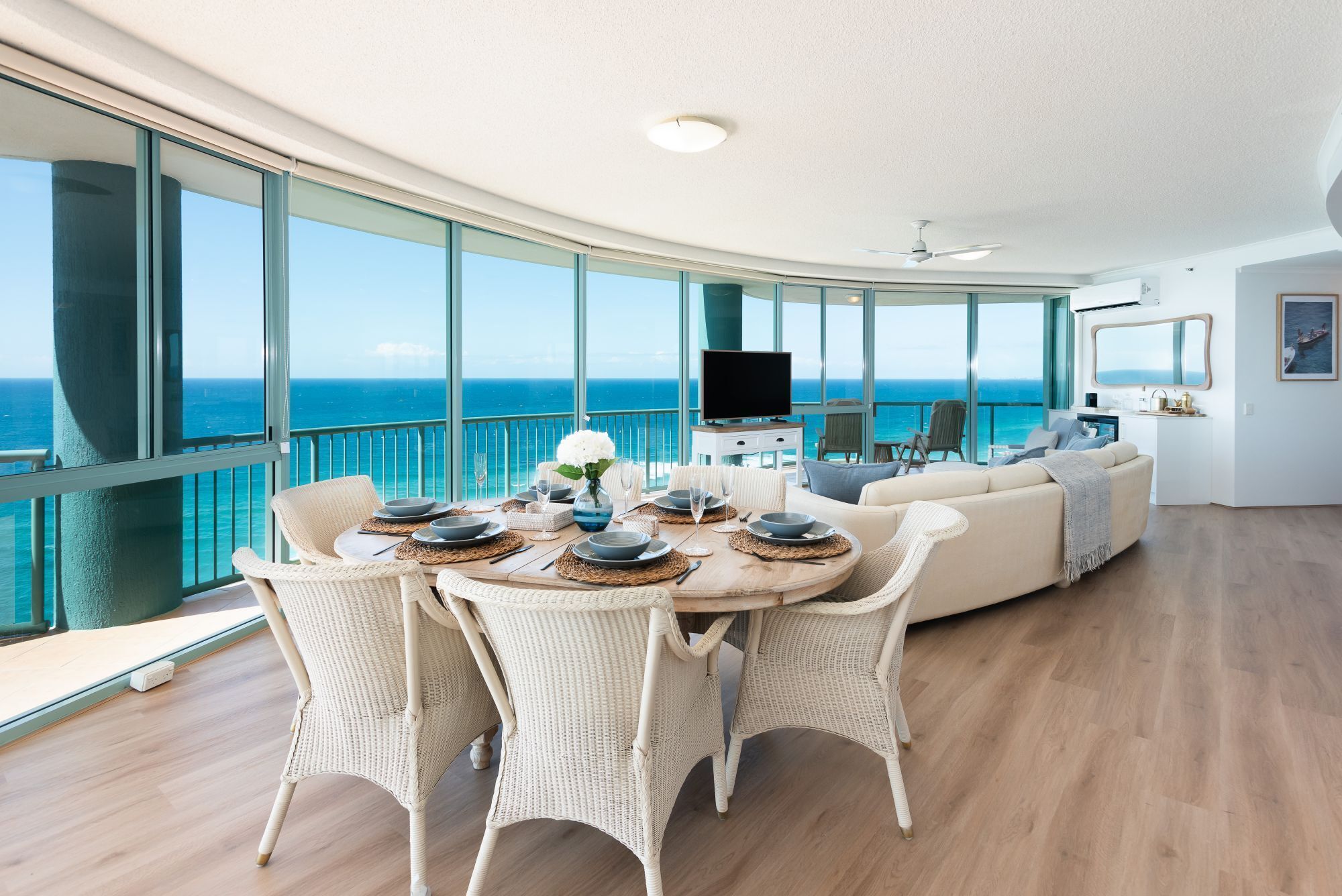 Uma sala de estar moderna com uma janela ampla com vista para o oceano, uma mesa de jantar redonda posta para quatro pessoas e uma área de estar confortável com um sofá e cadeiras.