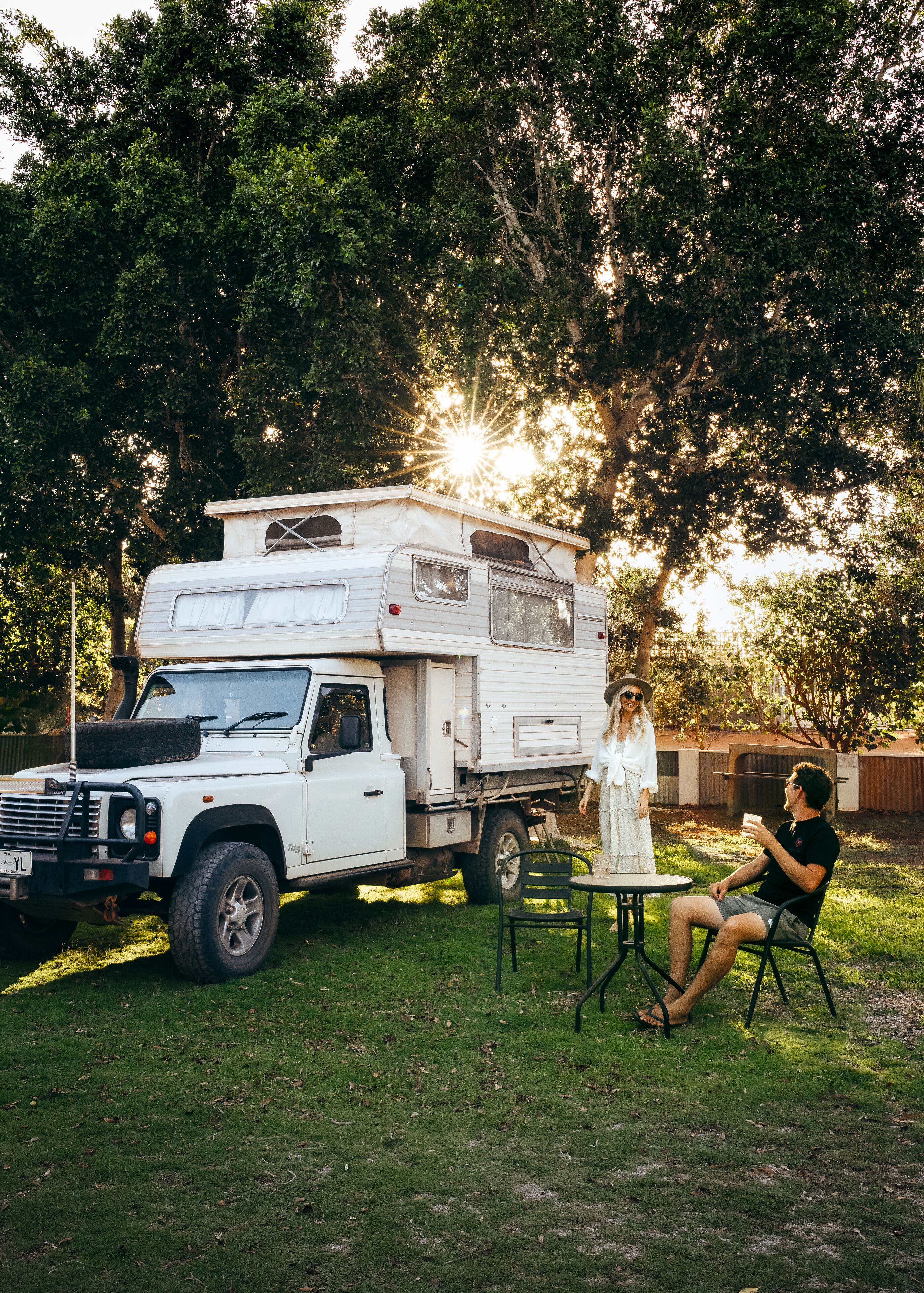 Uma carrinha camper branca estacionada na relva, com uma mesa e cadeiras por perto.