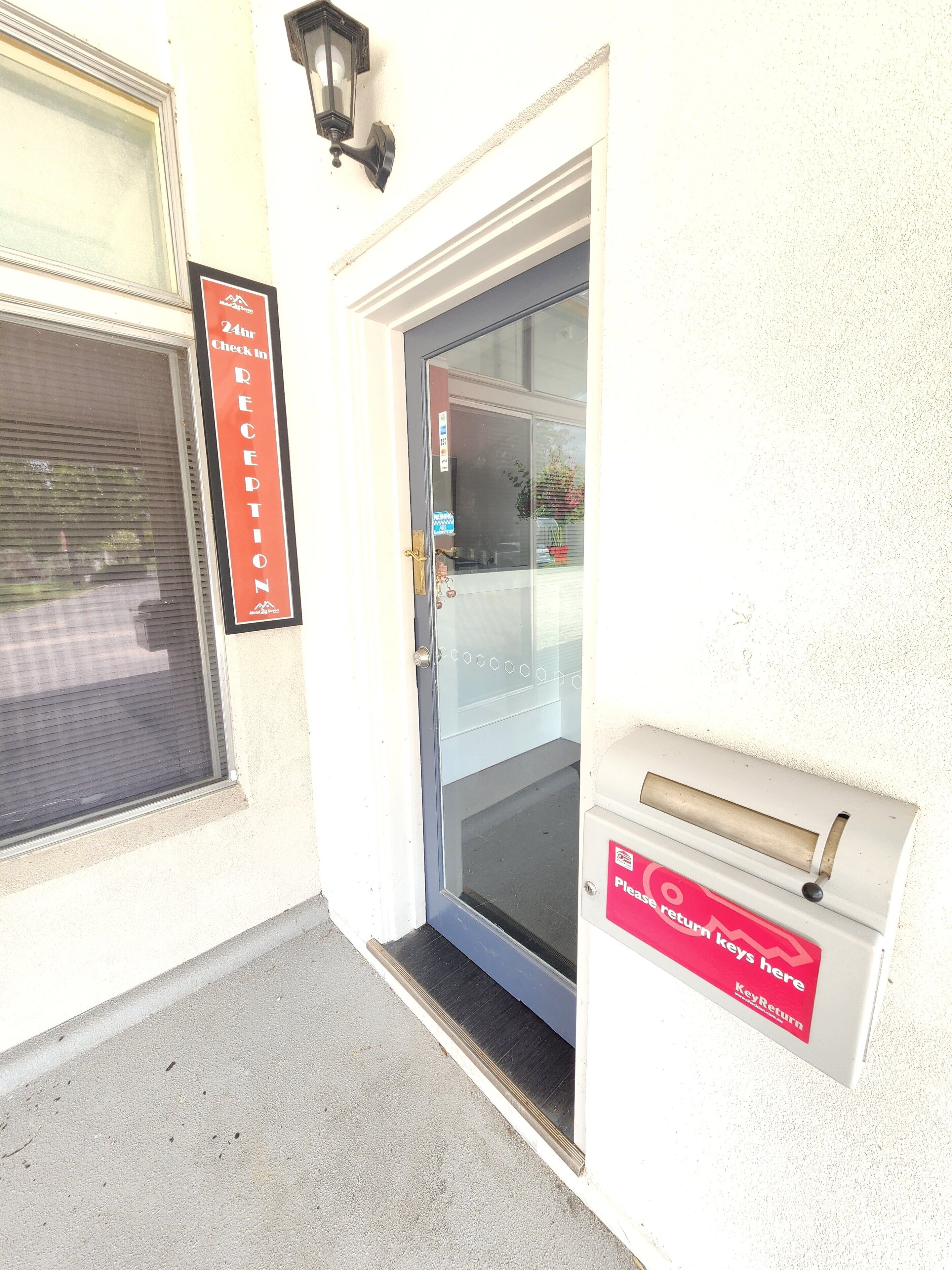 Entrada de hotel com placa de recepção, porta de vidro e caixa de correio com indicação de devolução de chaves.