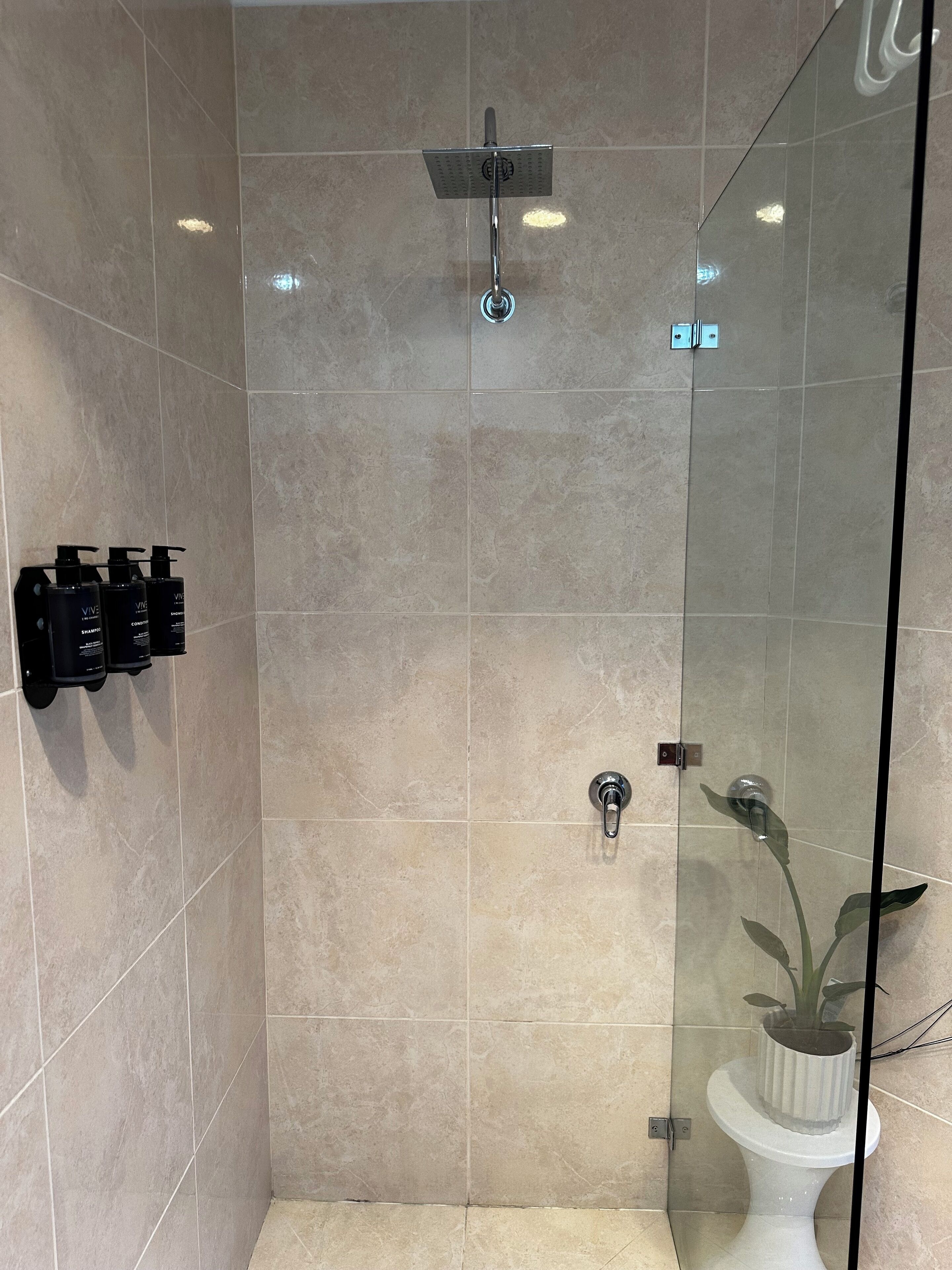 Uma área de chuveiro moderna, com box de vidro, chuveiro manual e dispensador de shampoo e condicionador fixado na parede.