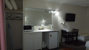 Quarto de hotel compacto com cozinha compacta, escrivaninha com micro-ondas, mini geladeira, cadeira e TV fixada na parede.