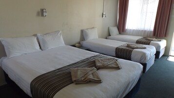 Quarto de hotel com três camas, cada uma com roupa de cama branca e manta listrada. Há dois interruptores de luz fixados na parede e uma janela com cortinas.