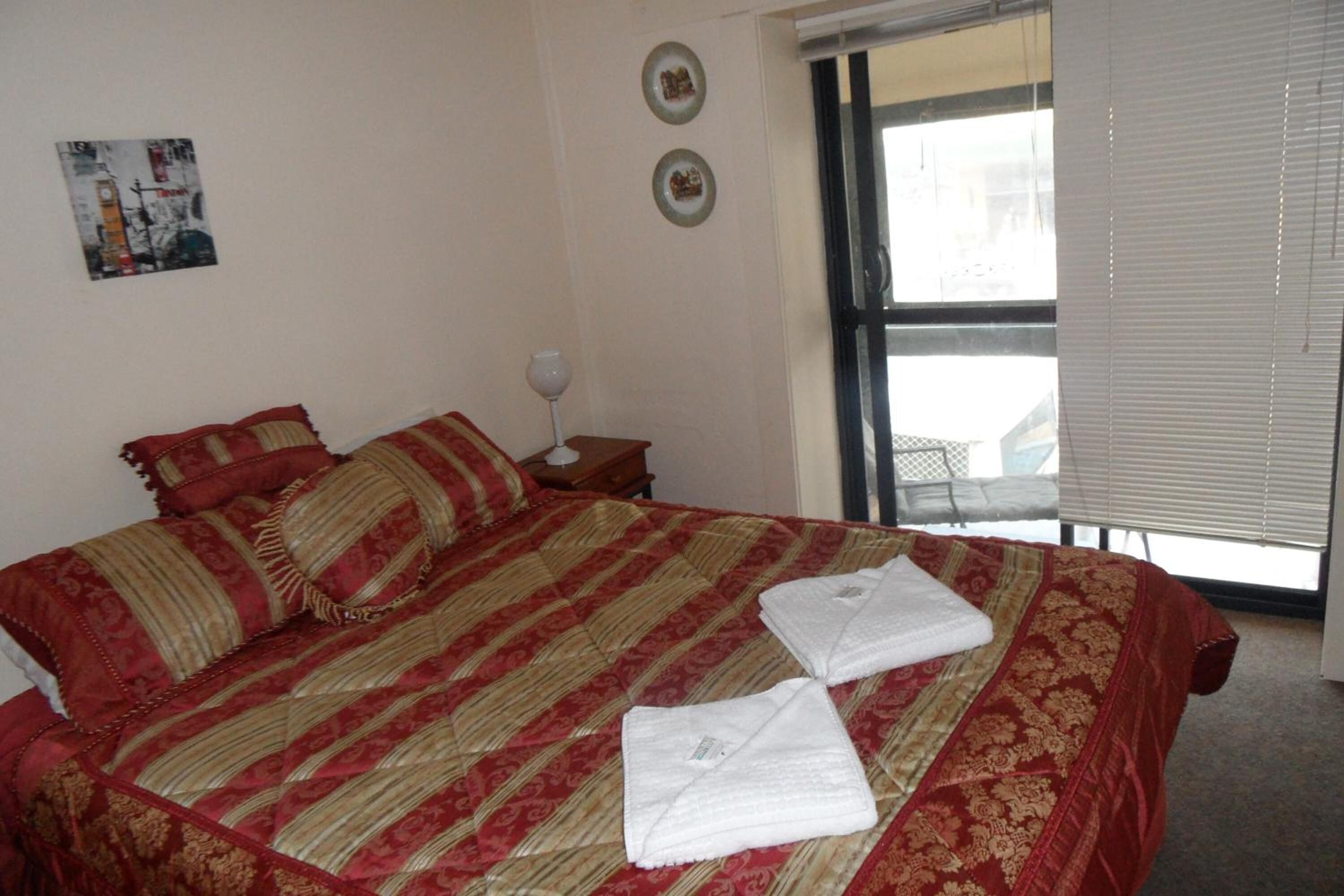 Quarto de hotel com uma cama grande, duas almofadas, mesa de cabeceira com abajur e janela com persianas.