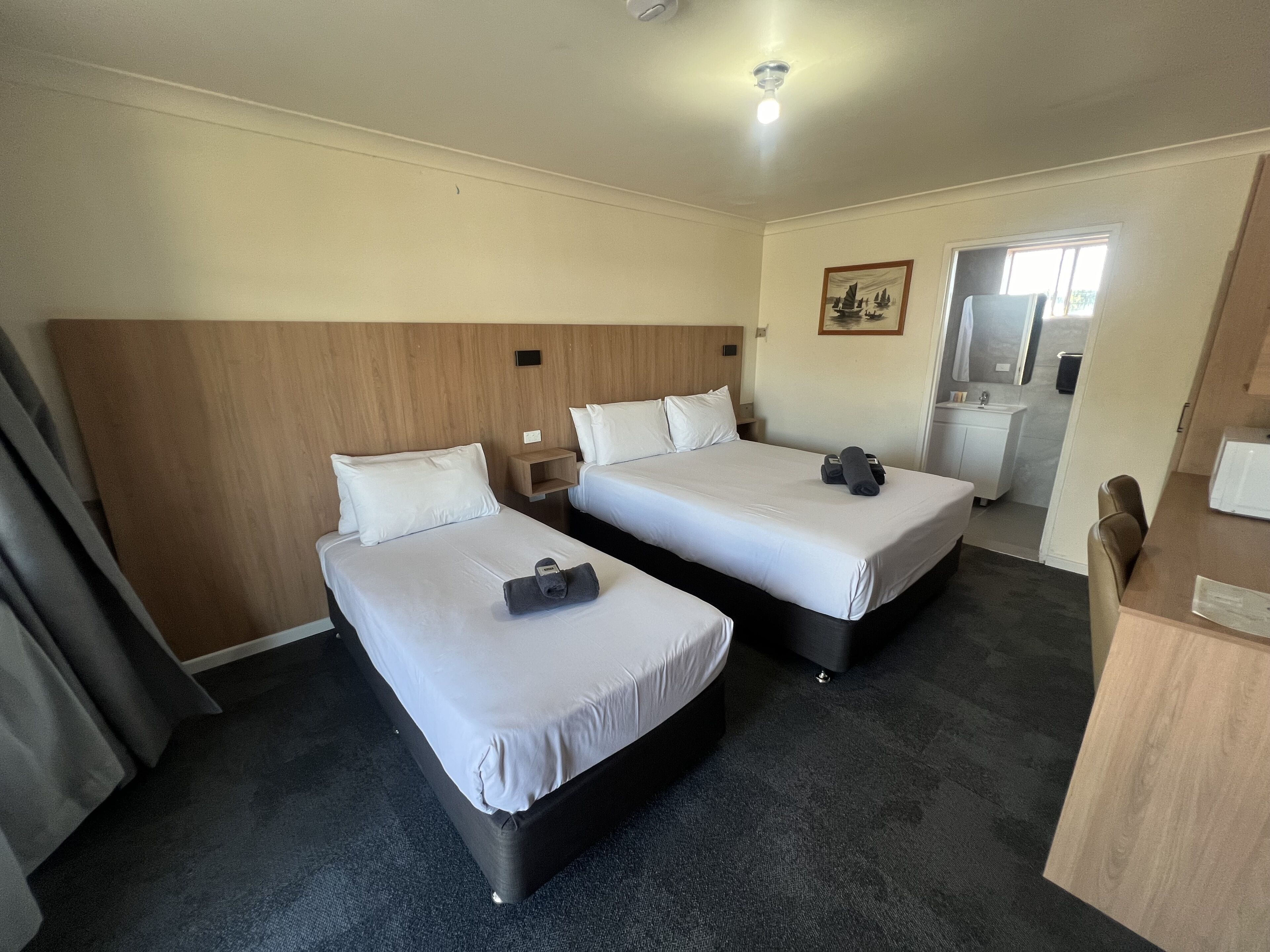 Quarto de hotel com duas camas, cabeceira de madeira, mesa de cabeceira pequena, espelho e banheiro visível por uma porta aberta.