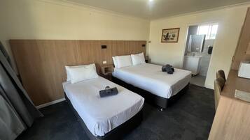 Quarto de hotel com duas camas, cabeceira de madeira, mesa de cabeceira pequena, espelho e banheiro visível por uma porta aberta.