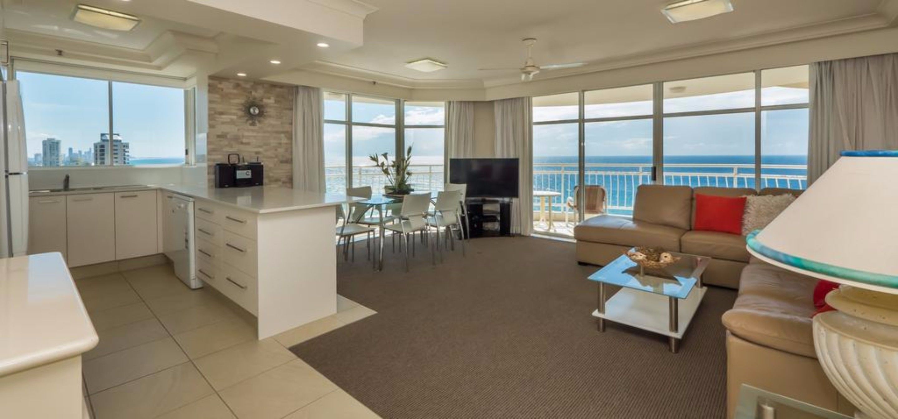 Sala de estar moderna com sofá, mesa de centro e televisão. Possui uma varanda com vista para o oceano e para a paisagem urbana.