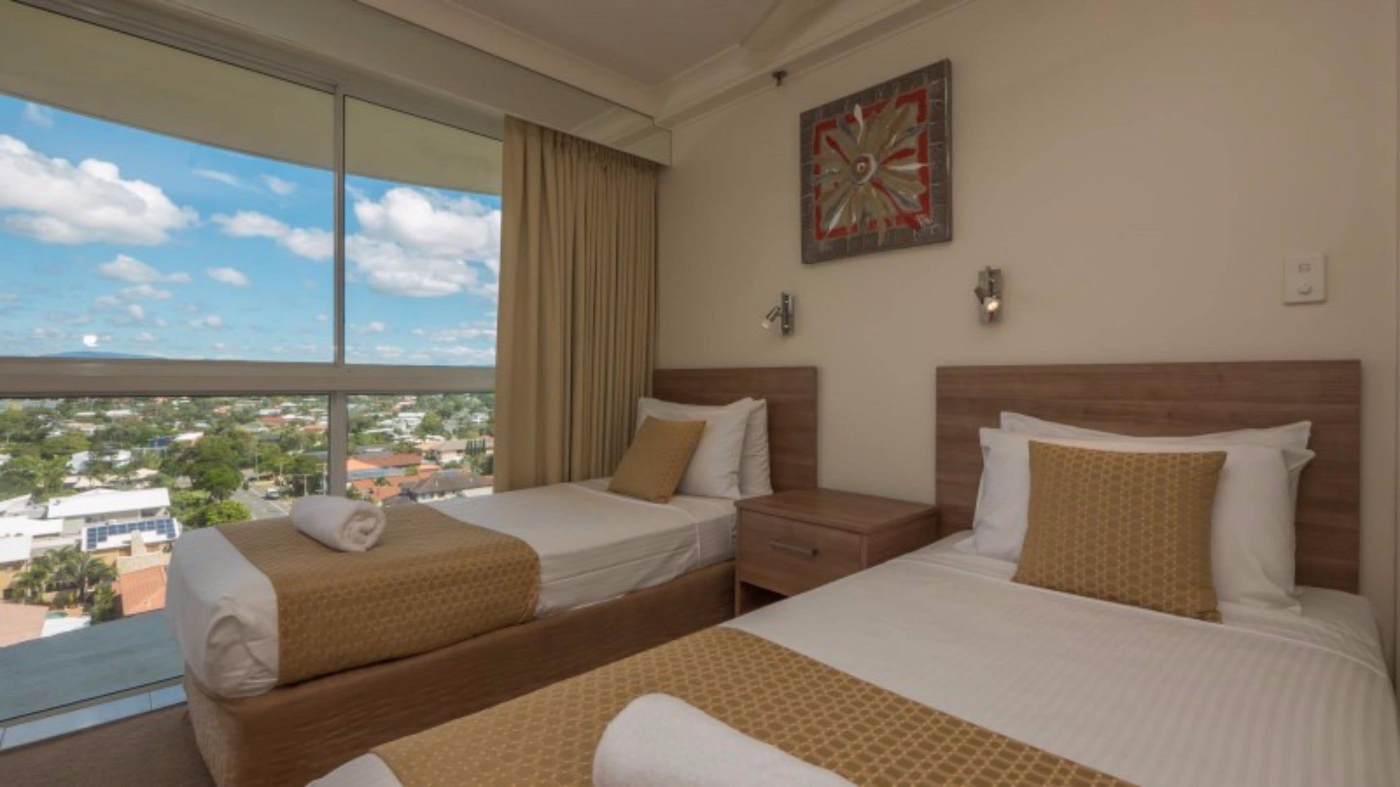 Quarto de hotel com duas camas, uma janela grande com vista para a cidade, um alvo de dardos na parede e uma mesa de cabeceira com abajur.