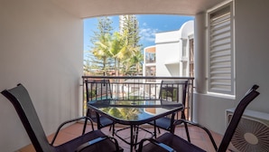 Uma varanda com mesa e cadeiras de vidro, um edifício branco ao fundo e vista para árvores e um céu claro.