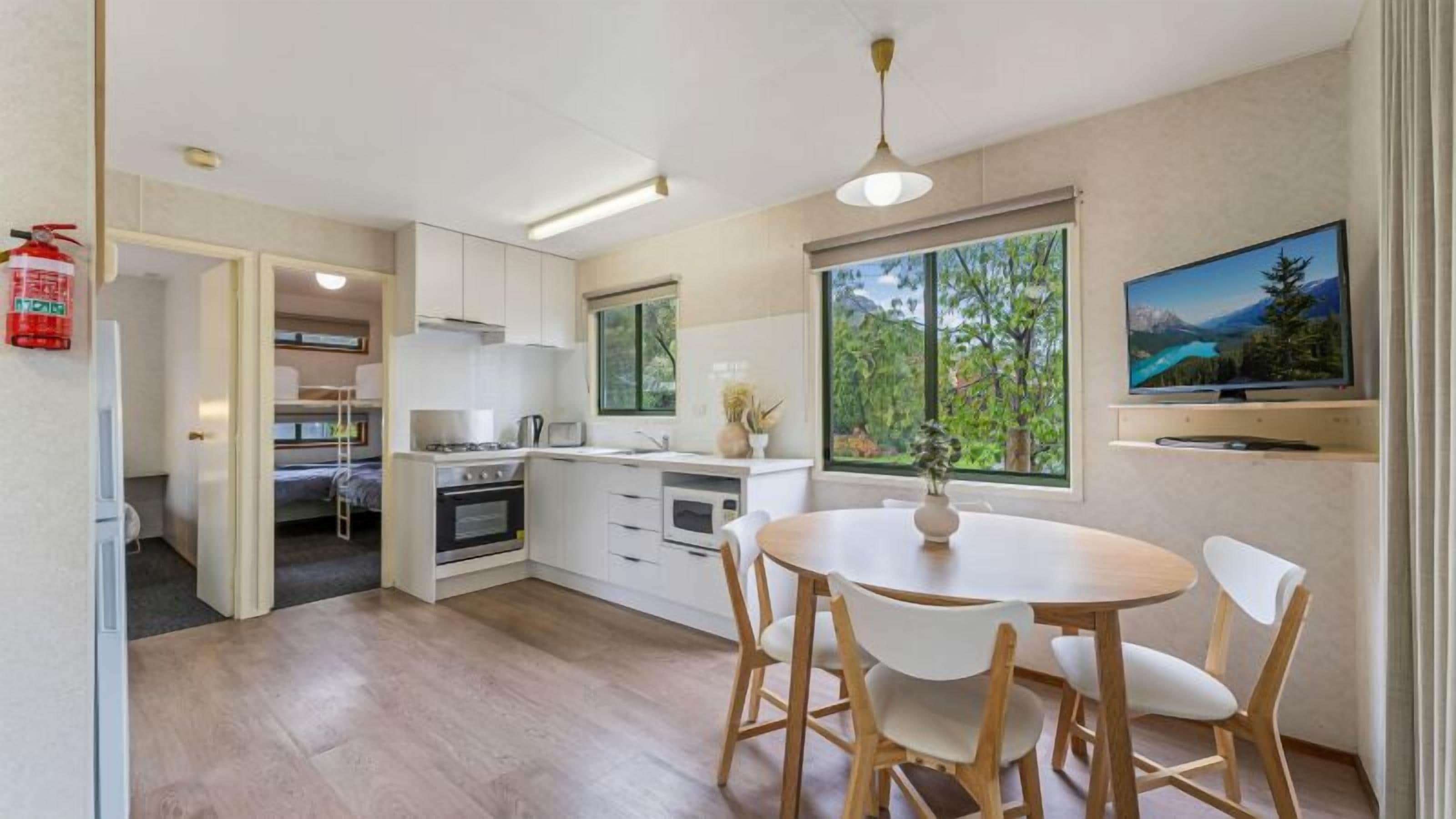 Uma cozinha moderna com armários brancos, um espaço de refeições com mesa redonda e cadeiras, e uma grande janela com vista para árvores e montanhas.