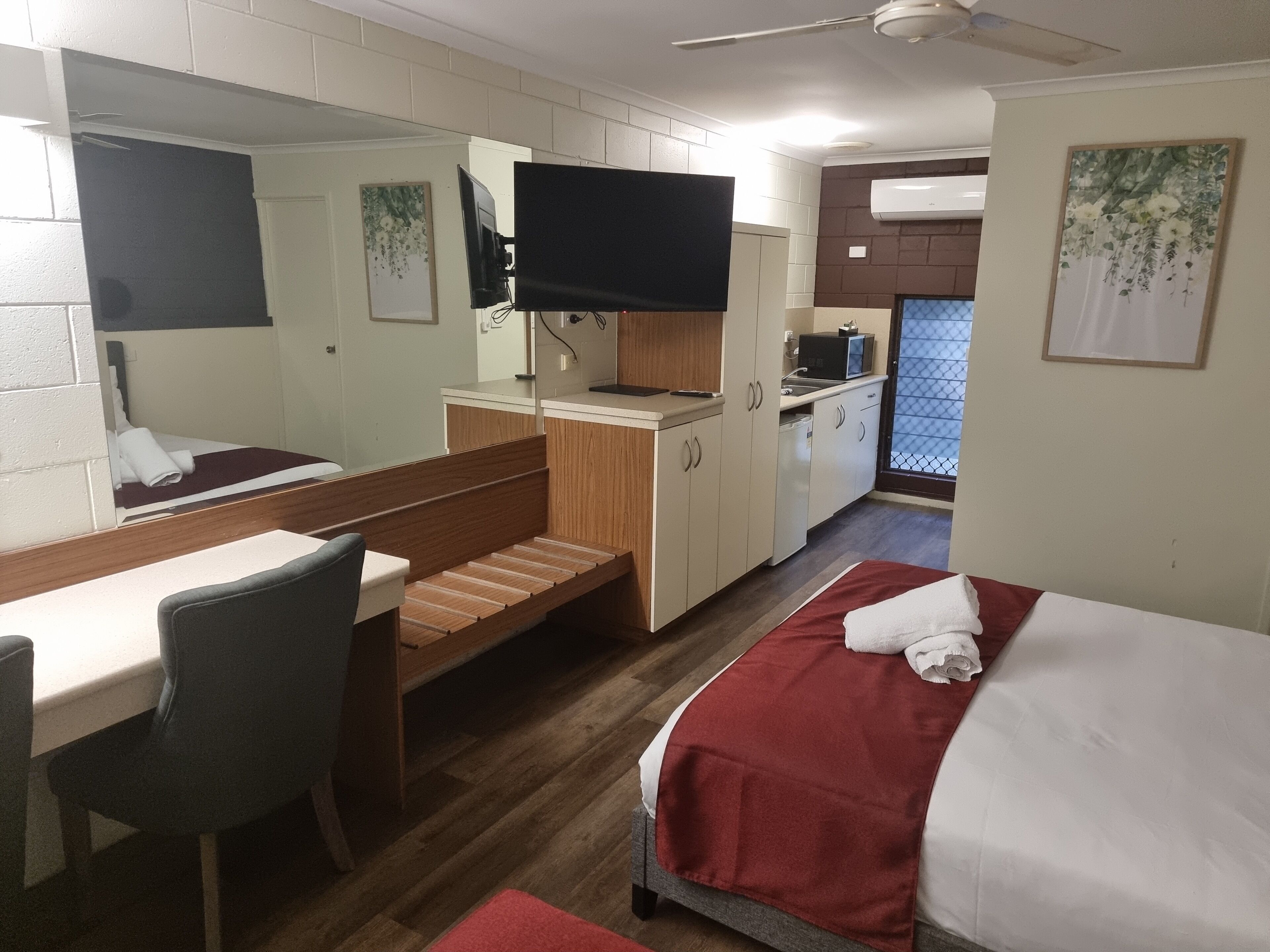 Quarto de hotel com uma cama, escrivaninha, cadeira, televisão e cozinha compacta.