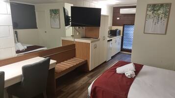 Quarto de hotel com uma cama, escrivaninha, cadeira, televisão e cozinha compacta.