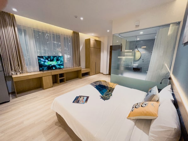 Quarto de hotel moderno com uma cama grande, televisão em prateleira embutida, banheiro com paredes de vidro e um guarda-roupa.