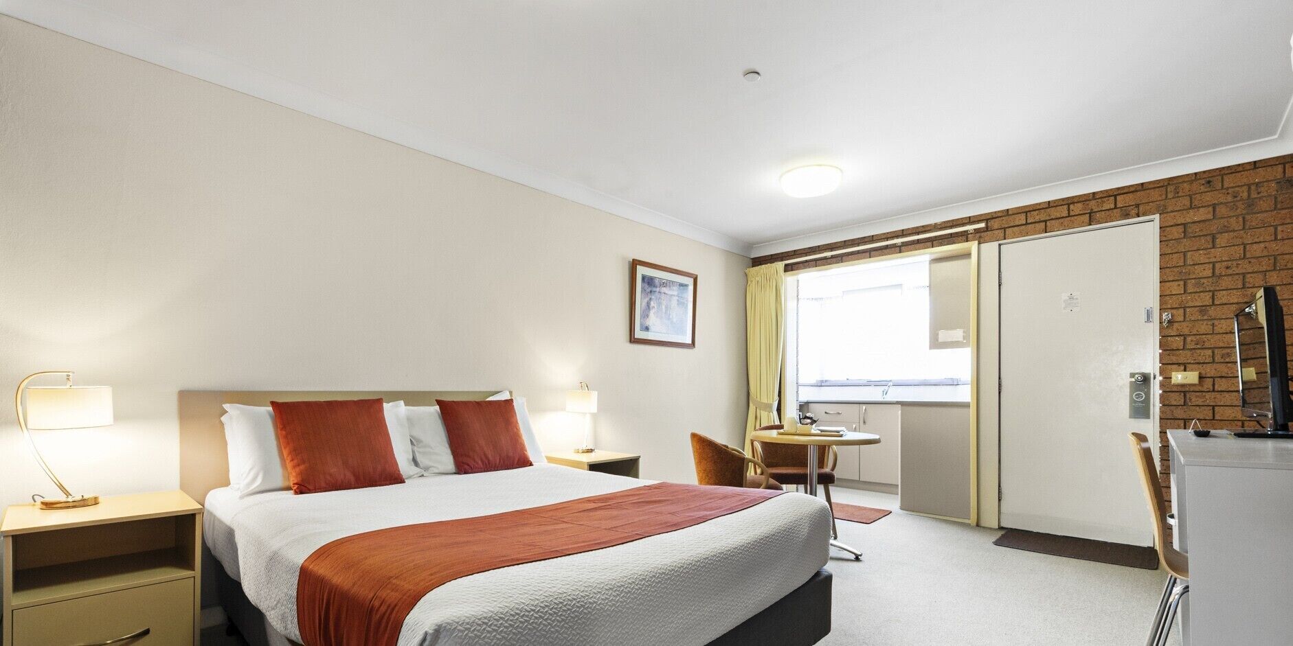Quarto de hotel com cama, mesas de cabeceira, escrivaninha, cadeira, mesinha e um frigorífico.