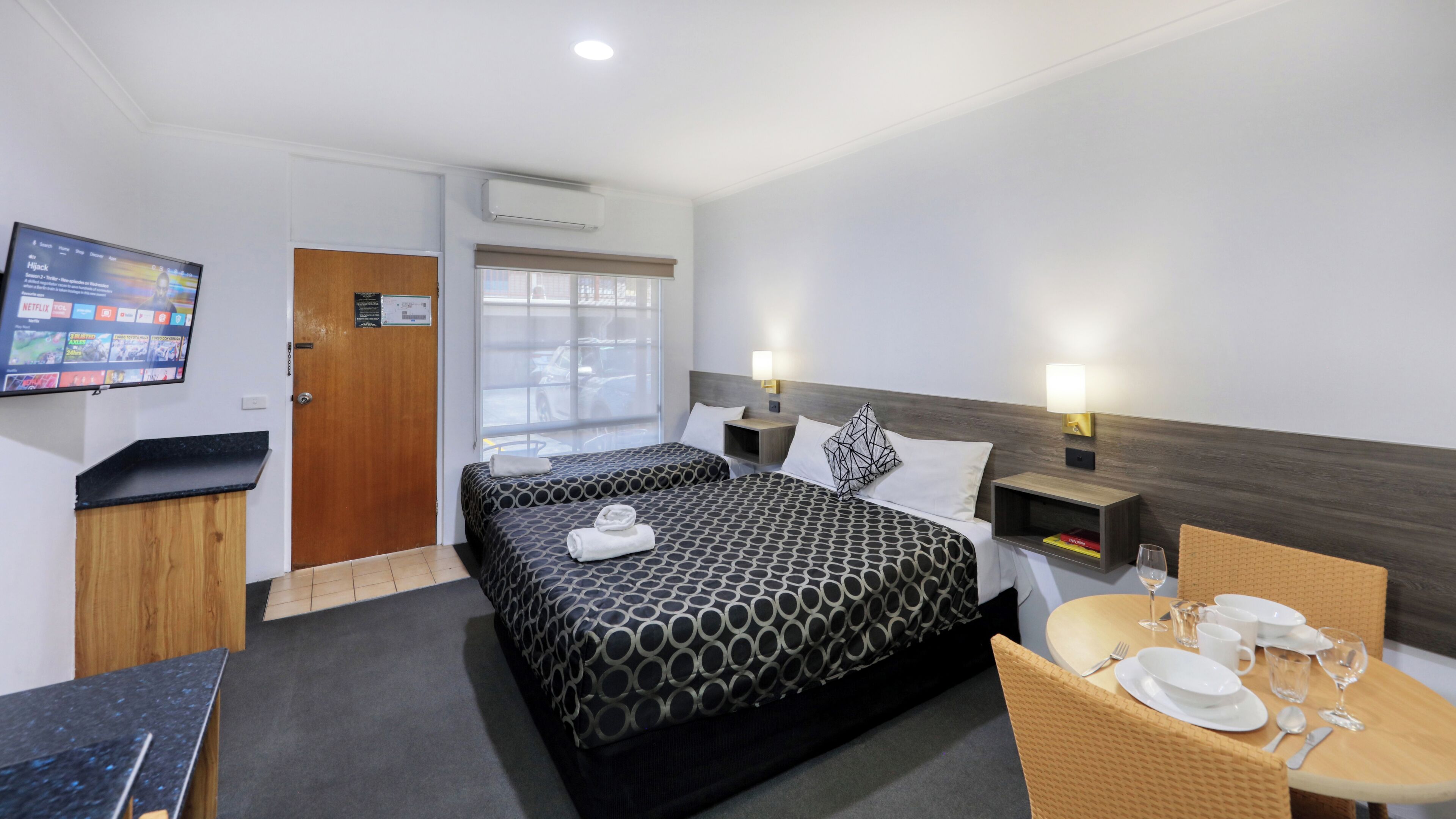 Quarto de hotel com duas camas, televisão, escrivaninha e área de refeições com mesa e cadeiras.