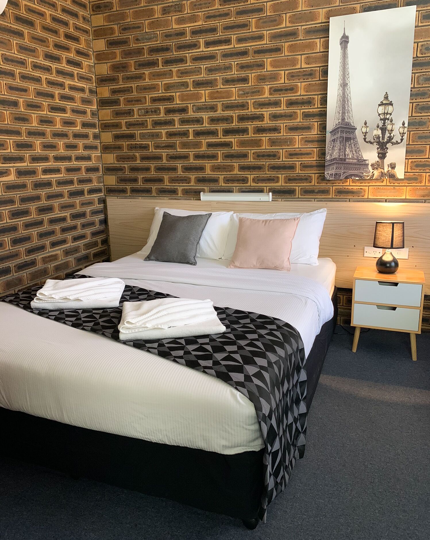 Quarto de hotel com parede de tijolos aparentes, cama com colcha de padrão geométrico e mesa de cabeceira com abajur.