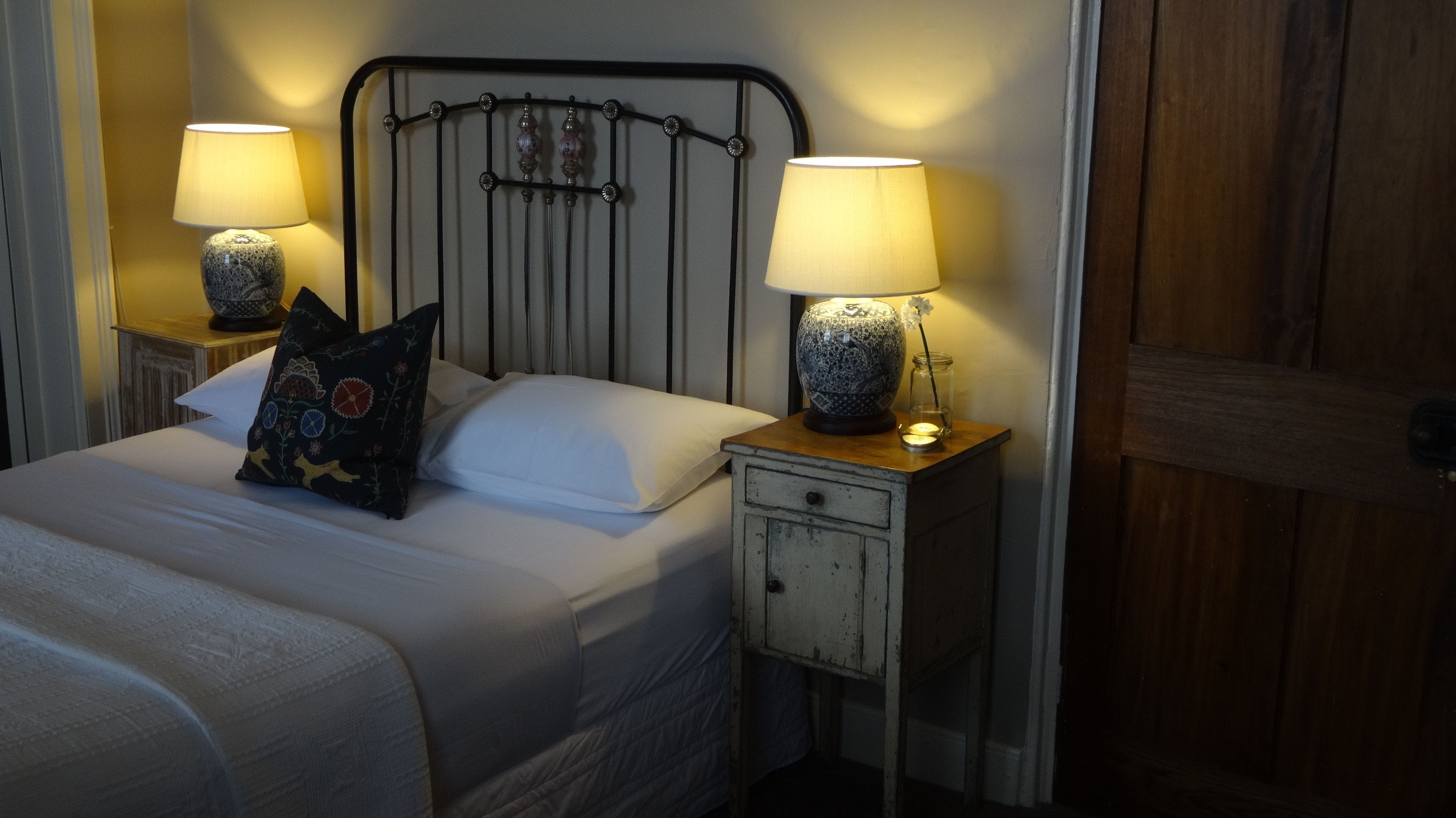 Cama bem arrumada com lençóis brancos, um travesseiro decorativo, duas mesas de cabeceira com luminárias e uma porta de madeira ao fundo.