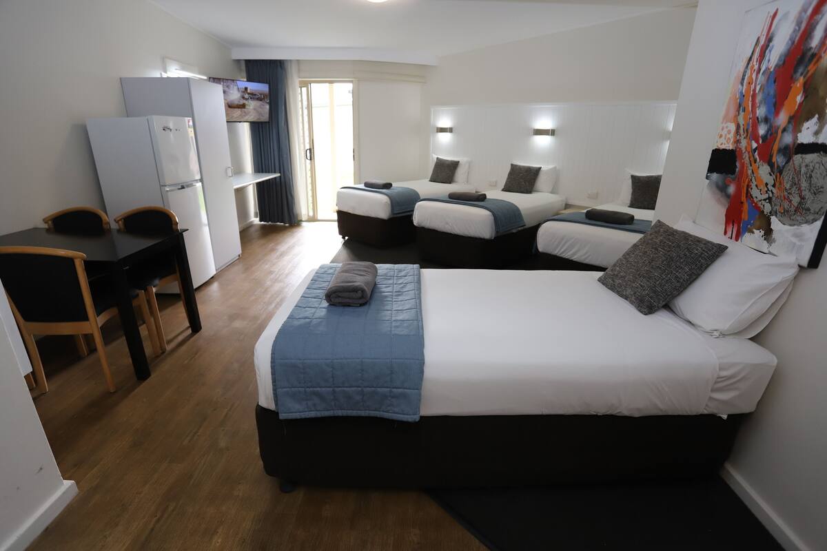 Quarto de hotel com cama, duas mesas de cabeceira, escrivaninha com cadeiras, geladeira e televisão.