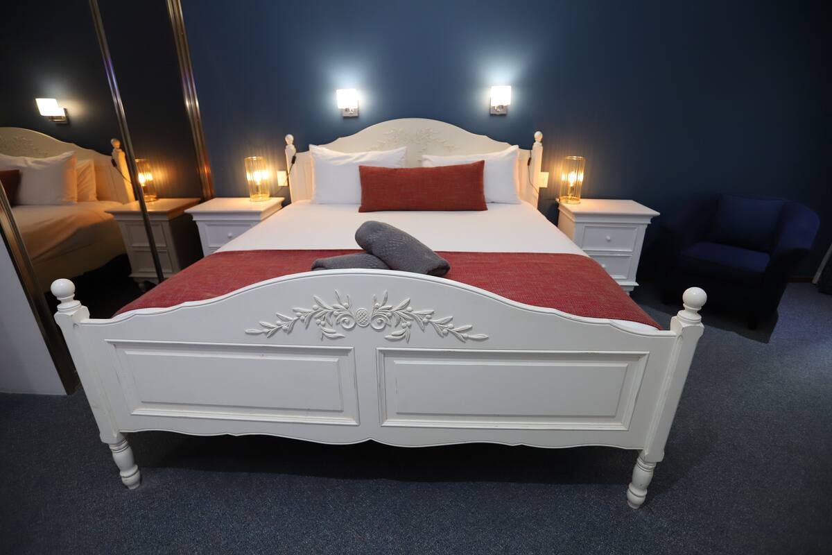 Uma cama branca com manta vermelha e almofadas, com mesas de cabeceira e abajures de cada lado, em quarto com paredes azul-escuro.