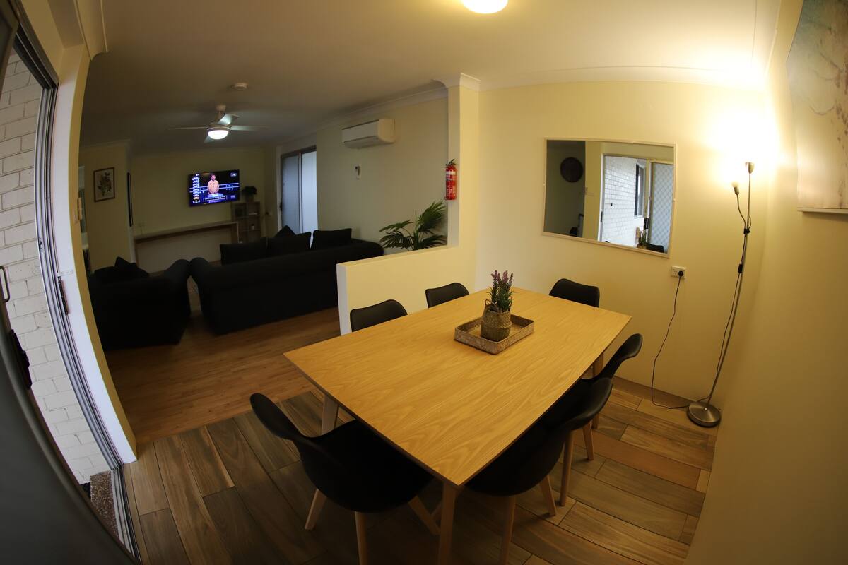 Uma área de refeições com uma mesa de madeira e cadeiras pretas, uma pequena planta em vaso e uma televisão ao fundo.