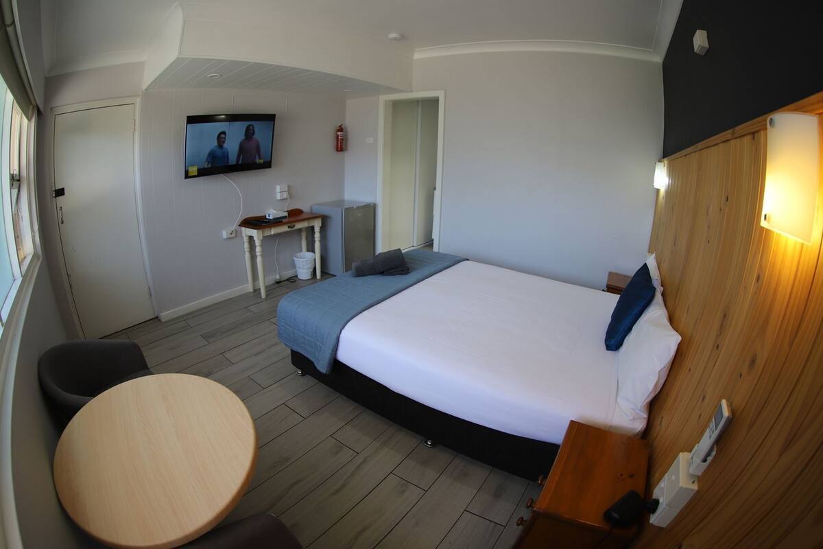 Quarto de hotel com cama, pequena escrivaninha, cadeira, mesa redonda e televisão na parede.