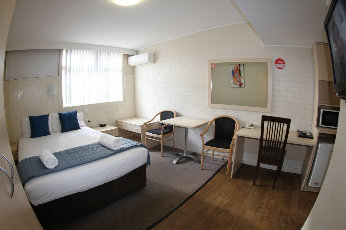 Quarto de hotel com cama, escrivaninha, cadeira e televisão.