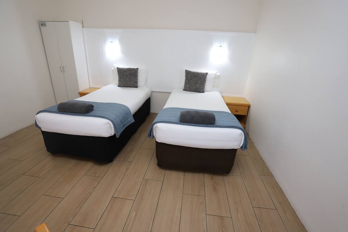 Quarto com duas camas, cada uma com travesseiro e cobertor, um guarda-roupa e uma mesa de cabeceira com luminária.