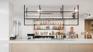 Uma área de bar com prateleiras cheias de garrafas e copos, um balcão com um caixa registradora e uma pessoa preparando bebidas.