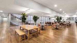 Uma sala de jantar espaçosa e moderna, com mesas e cadeiras de madeira, janelas amplas e plantas em vasos.