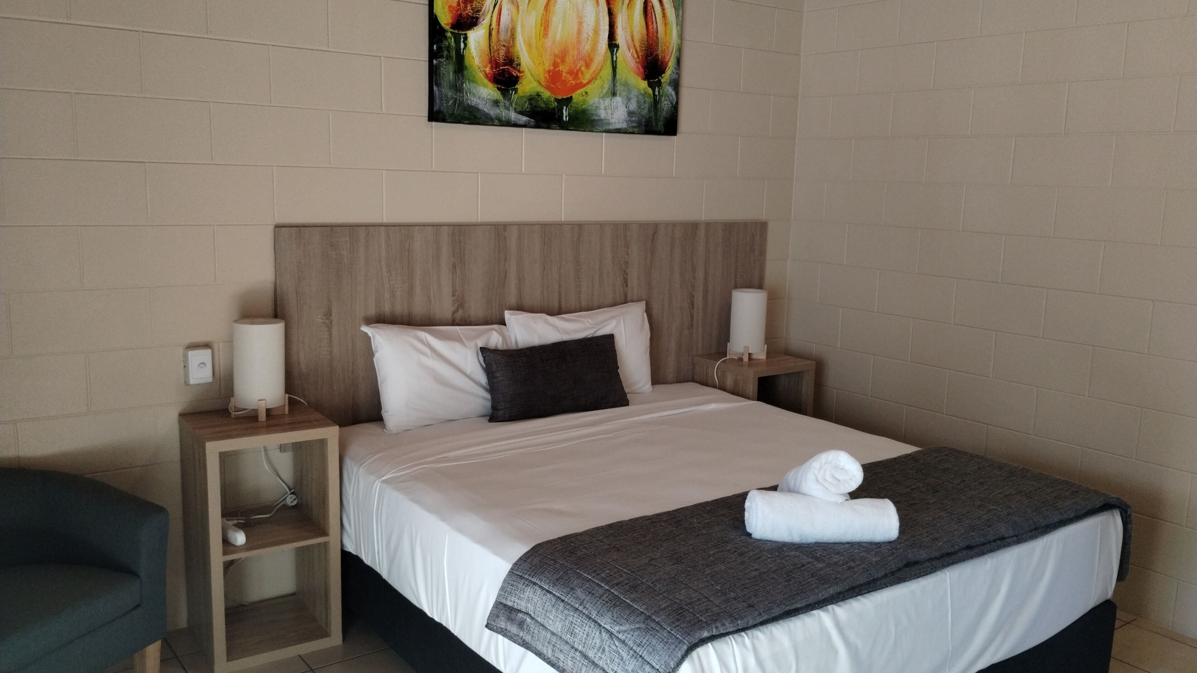 Quarto de hotel com uma cama, duas mesas de cabeceira, um criado-mudo, um quadro na parede e uma cadeira.