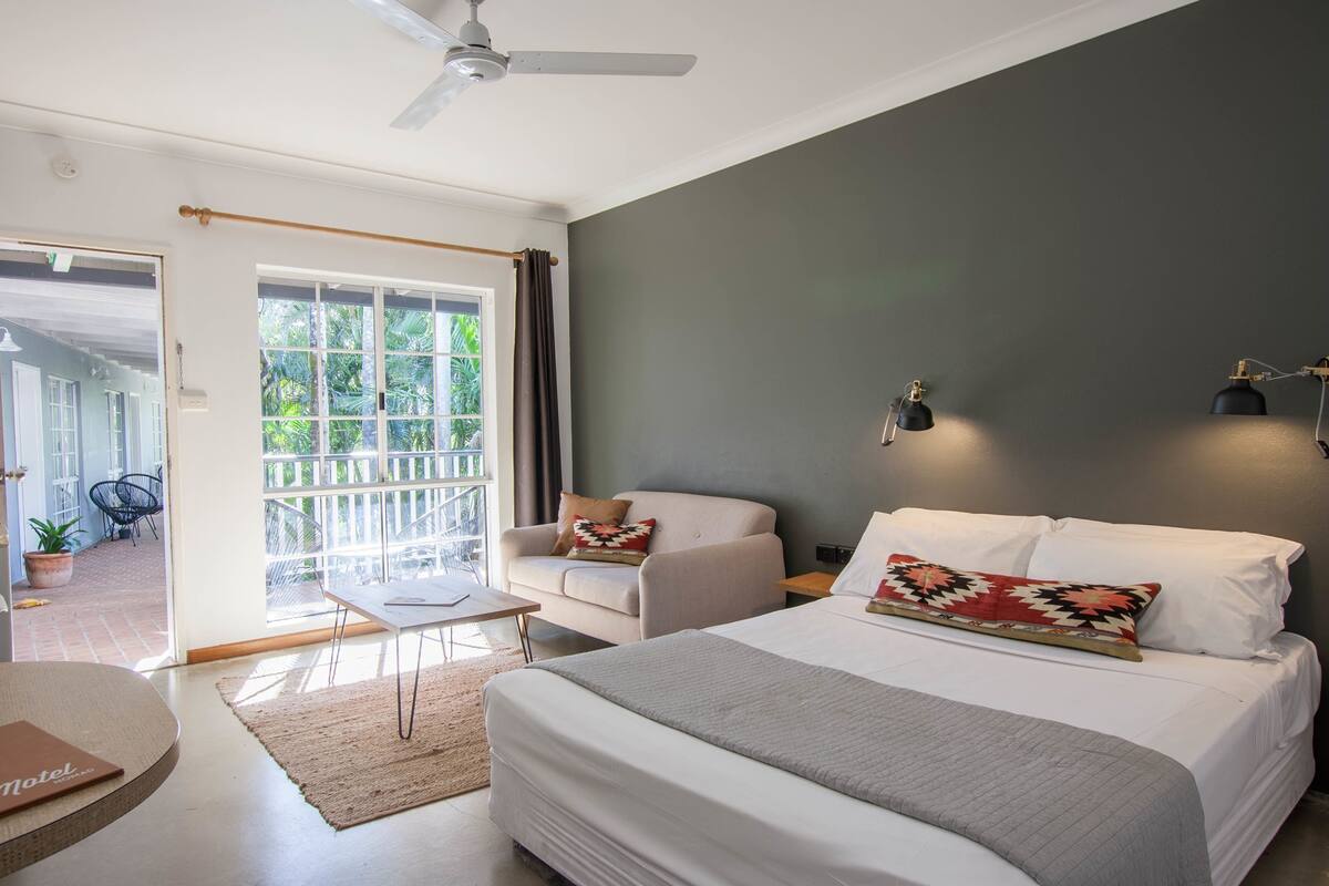 Quarto de hotel com cama, sofá, mesa pequena, ventoinha no teto e vista para o exterior.