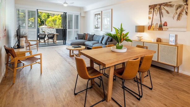 Uma sala de estar moderna com uma mesa de jantar de madeira, assentos confortáveis, um sofá e vista para uma área externa com plantas através das portas de vidro.