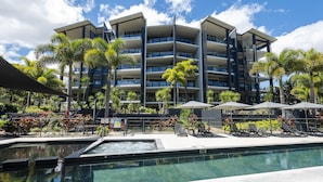 Um hotel moderno com área de piscina, incluindo espreguiçadeiras e guarda-sóis, cercado por palmeiras e um céu azul límpido.