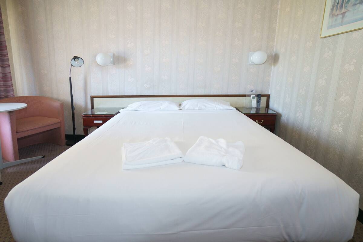 Cama bem arrumada com lençóis e toalhas brancos, mesa de cabeceira com abajur, e uma cadeira em quarto de hotel com papel de parede listrado.