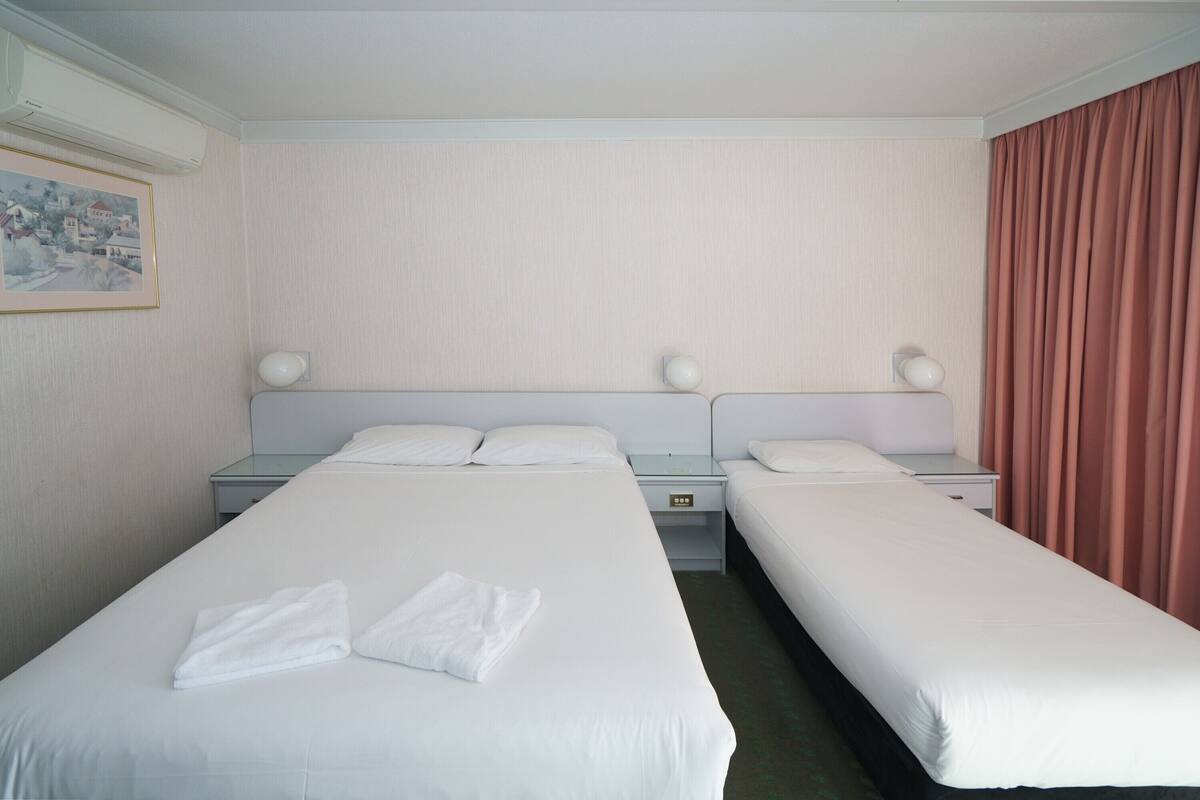 Quarto de hotel com duas camas de solteiro, mesa de cabeceira com abajur, quadro pendurado na parede e janela com cortinas.