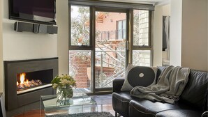 Uma sala de estar com lareira, um sofá, uma mesa de centro e uma janela grande com vista para um edifício de tijolos e uma árvore.