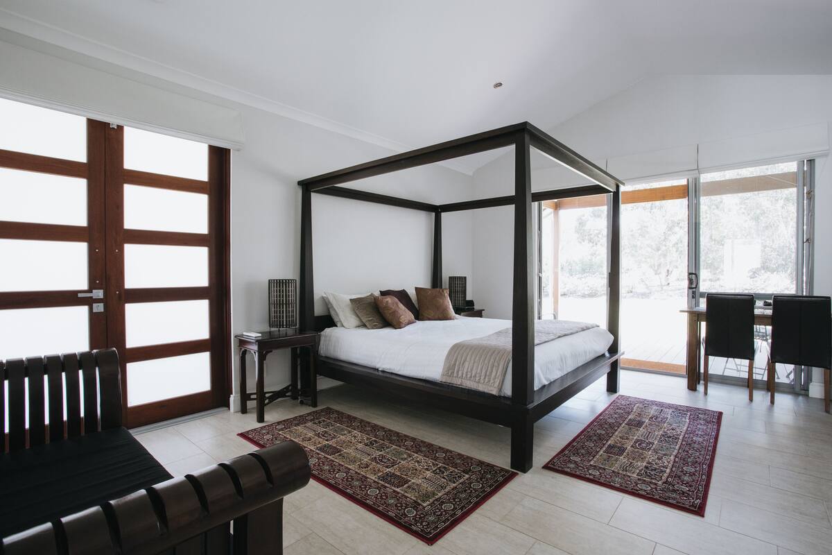 Uma cama com dossel e estrutura de madeira, uma escrivaninha com duas cadeiras e vista para o exterior através de amplas janelas.
