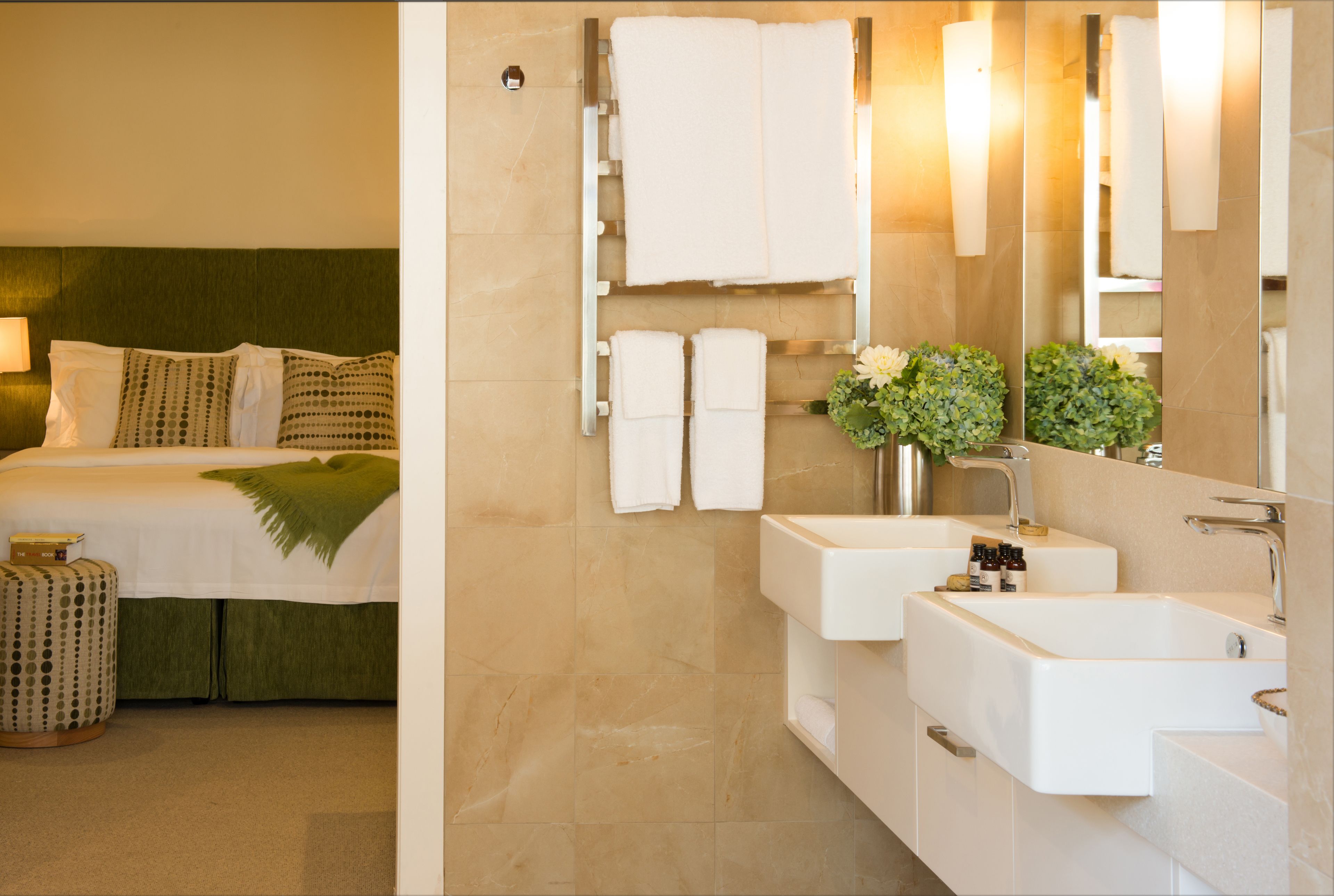 Quarto de hotel com cama, almofadas, mesa de cabeceira, manta verde, banheiro com duas pias, espelho e planta em vaso.