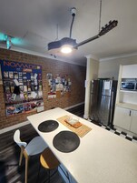 Uma cozinha com bancada branca, geladeira de aço inoxidável e micro-ondas. Há uma parede com um mural e uma placa com a inscrição 'Nomads'.